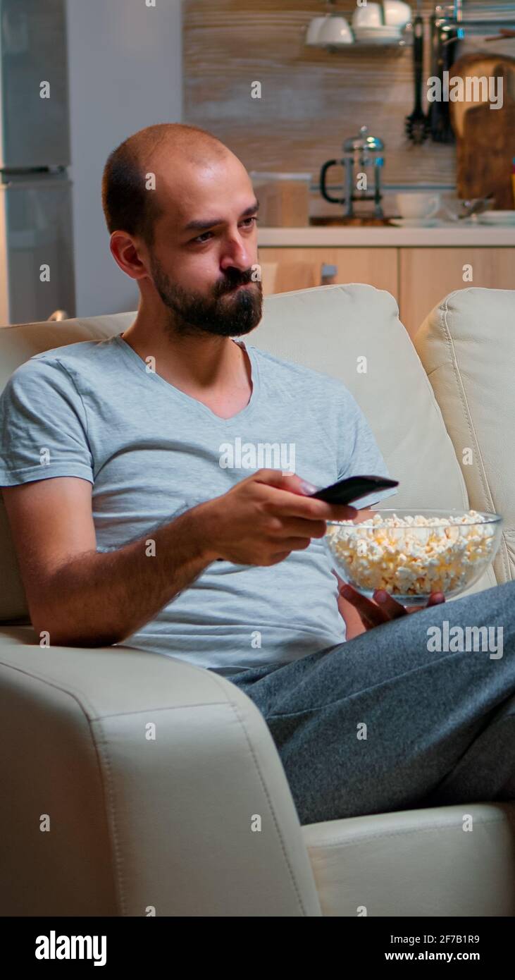 Bored man switching TV channels, holding popcorn in hads late at night ...