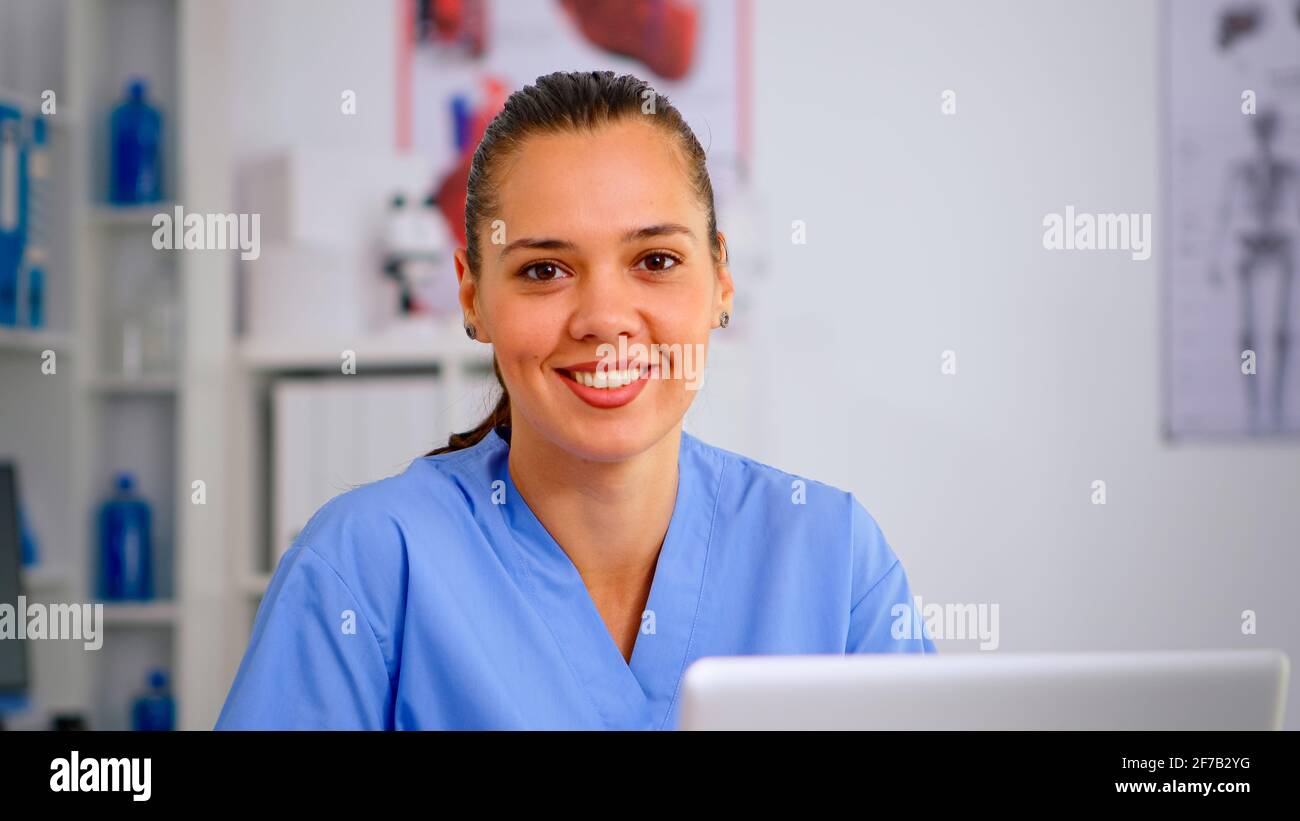Close up of young medical assistant typing at laptop and raising head ...