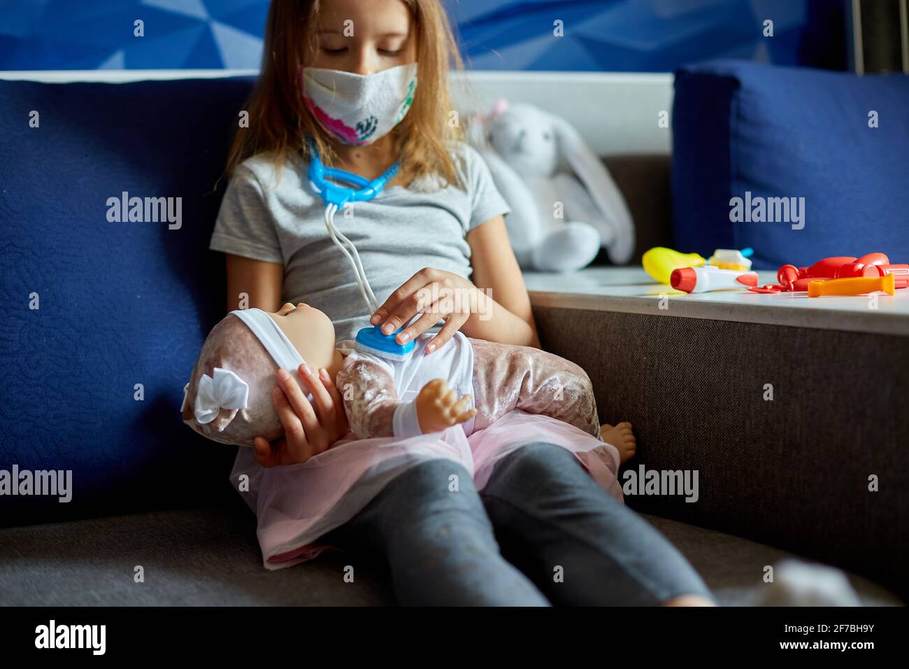 A little girl in medical mask sits on the sofa, plays a doctor, treats ...
