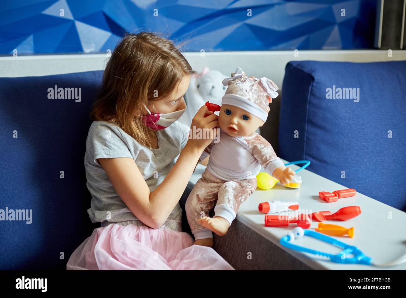 A little girl in medical mask sits on the sofa, plays a doctor, treats ...