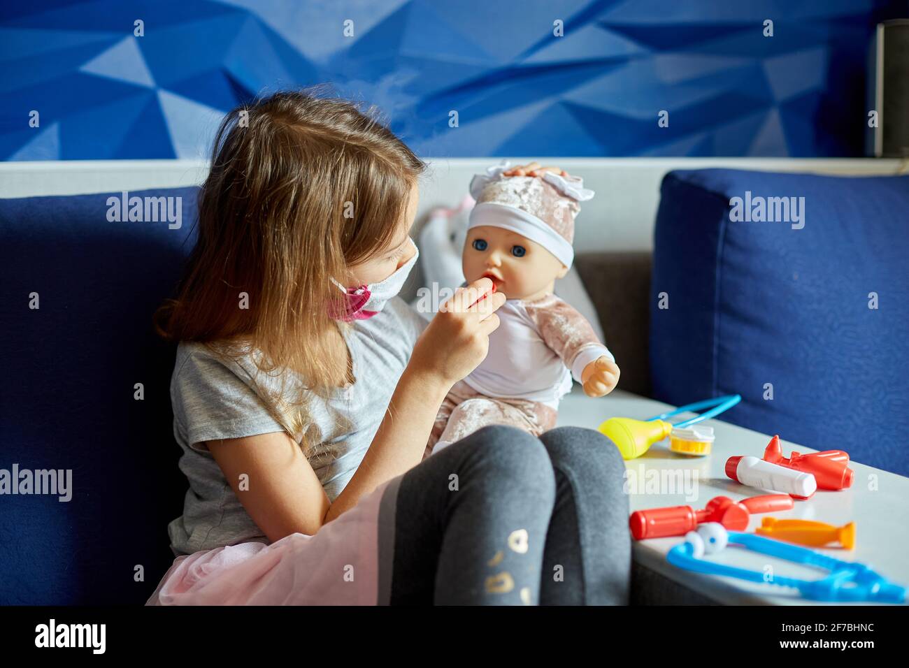 A little girl in medical mask sits on the sofa, plays a doctor, treats ...