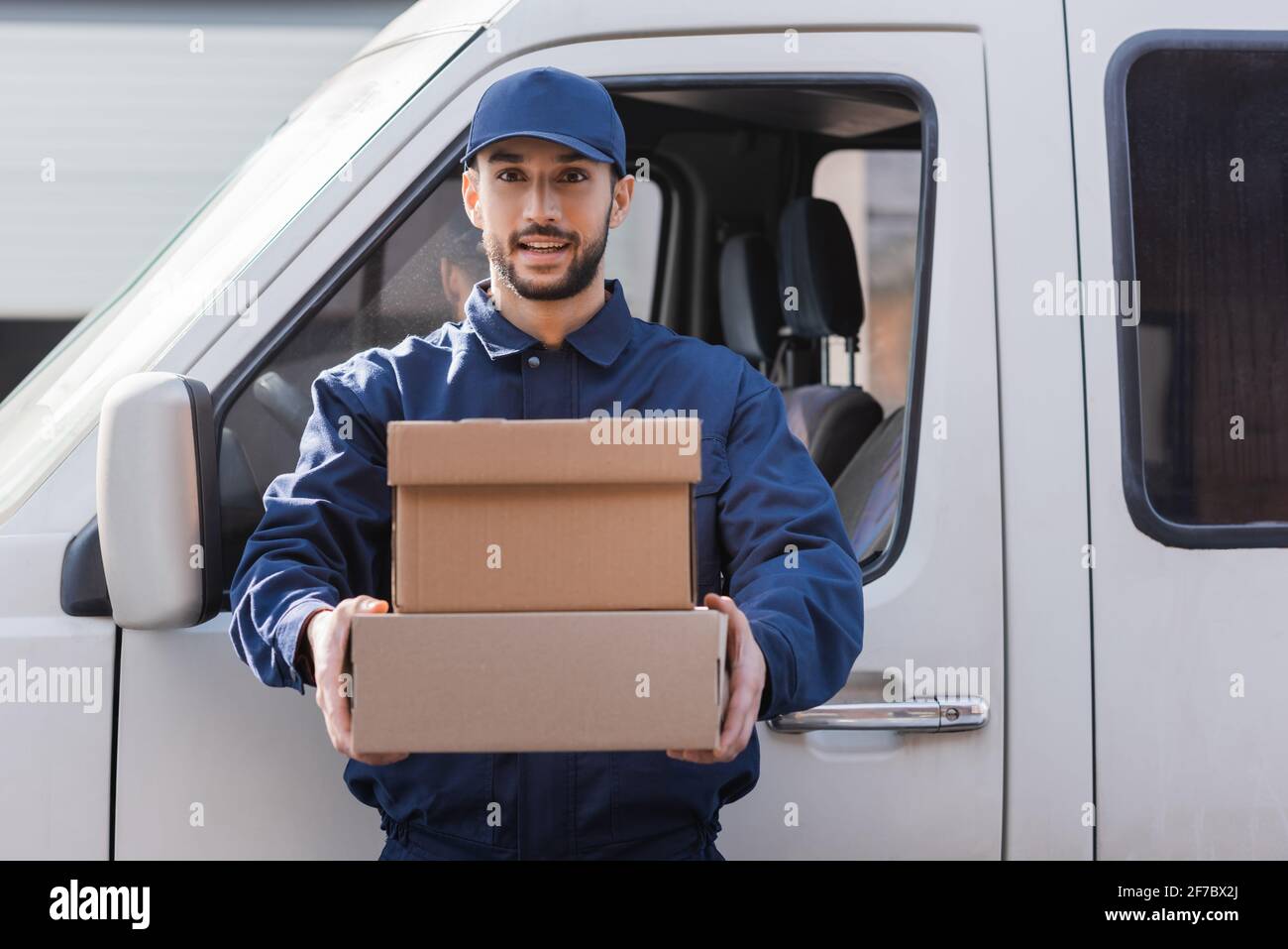 young arabian postman holding parcels and looking at camera near car ...