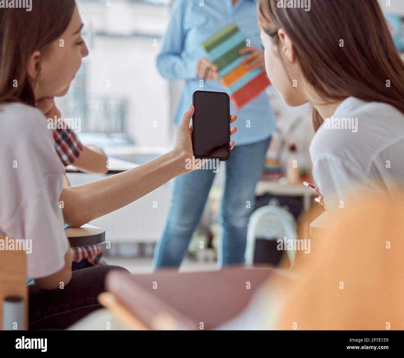 Close up girl shows smartphone to her classmate while sitting at desk ...
