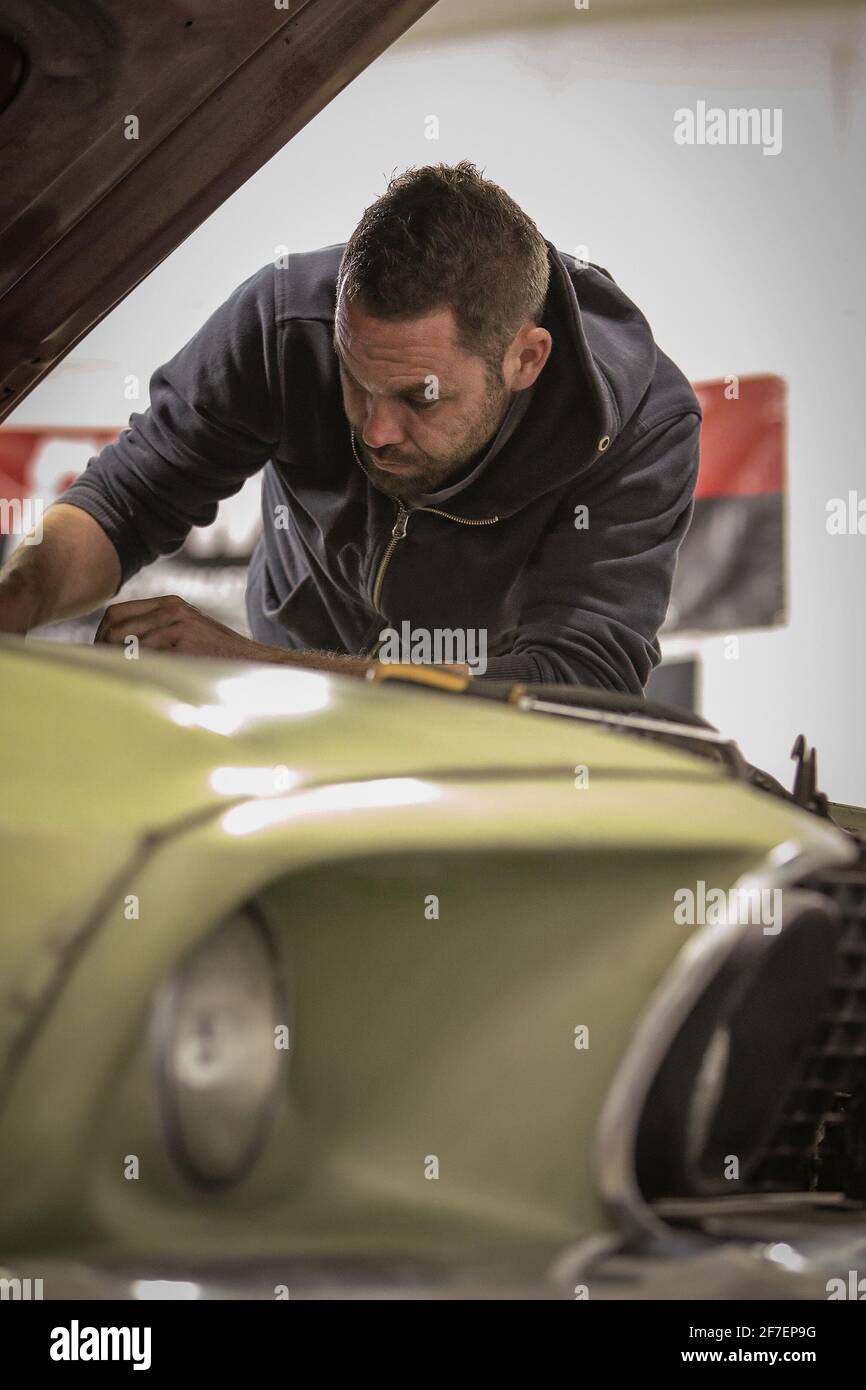 A young man is reaching under the hood of an old vintage car fixing the ...