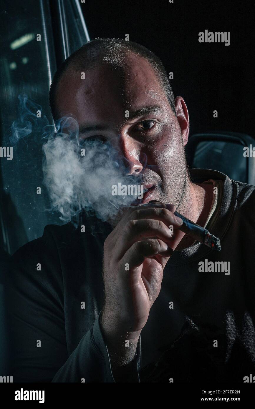 A young man with a serious expression is smoking a cigar in a dark ...