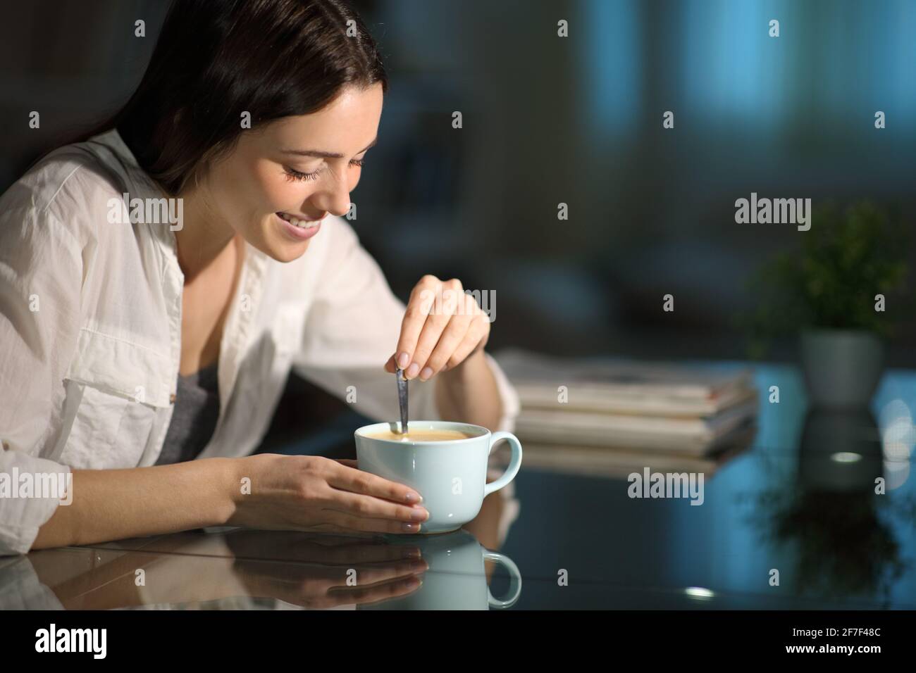 Happy woman stirring coffee sitting in the living room in the night at ...