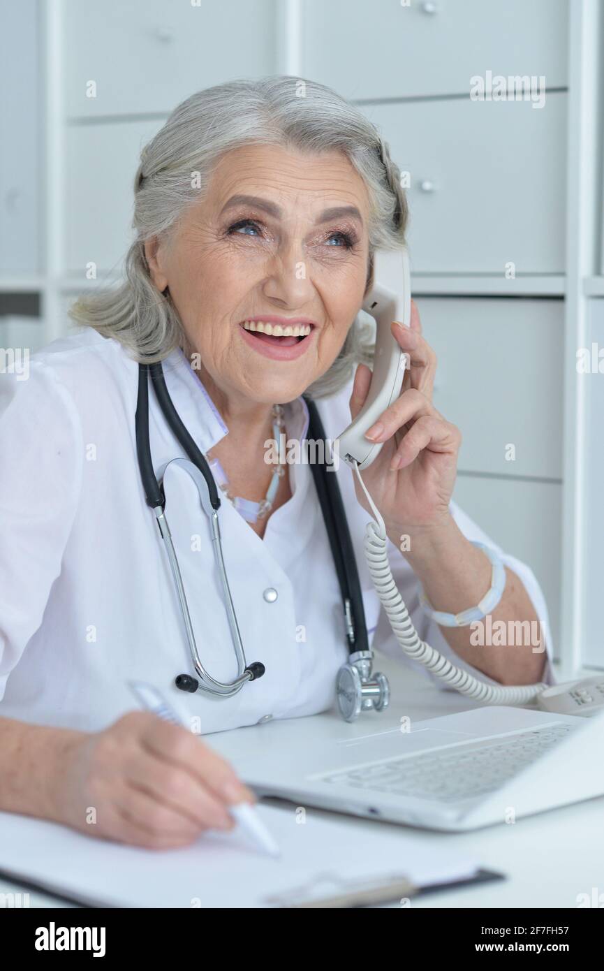 Female doctor talking on the phone while working at table with laptop ...