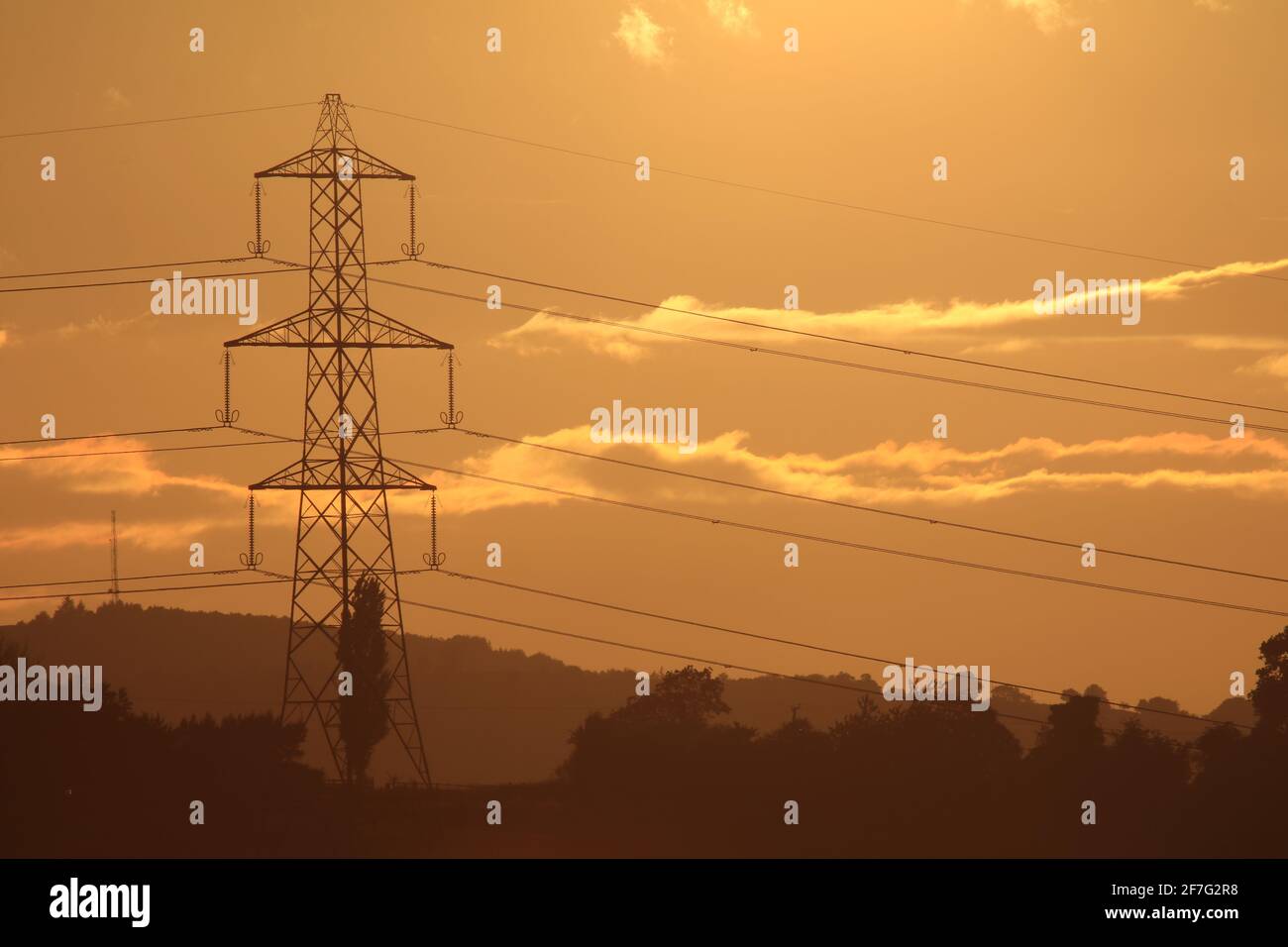 Electricity Pilon against a sunset sky and could, trees and hills in ...