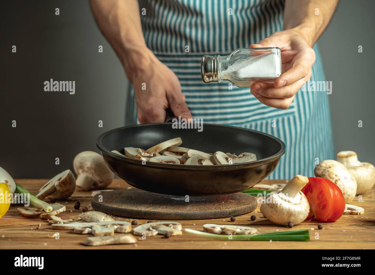 A chef in a blue apron is sprinkling salt on mushrooms in a frying pan ...