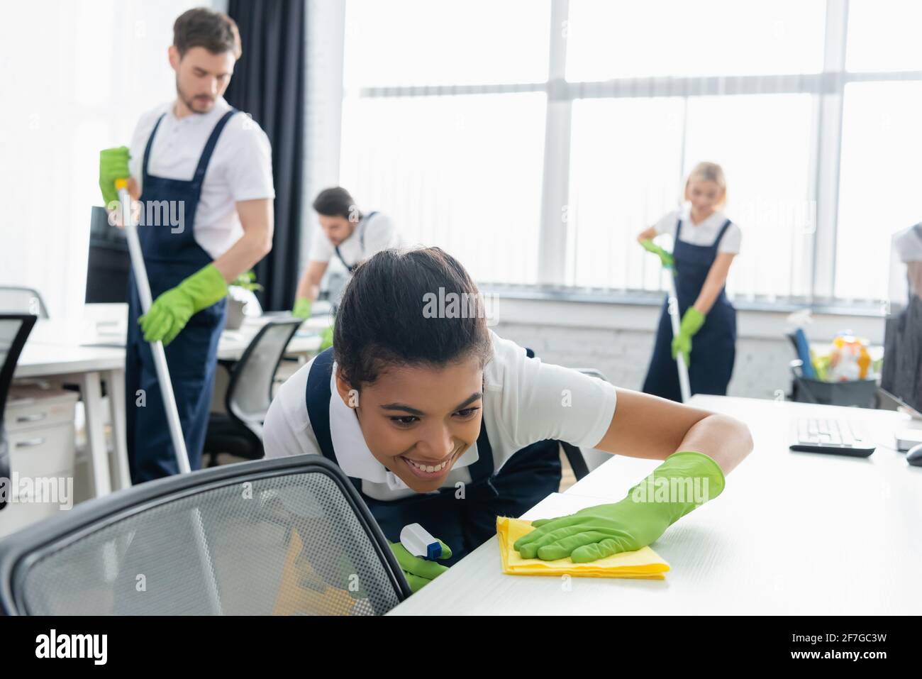 Smiling african american cleaner cleaning table with rag in office ...