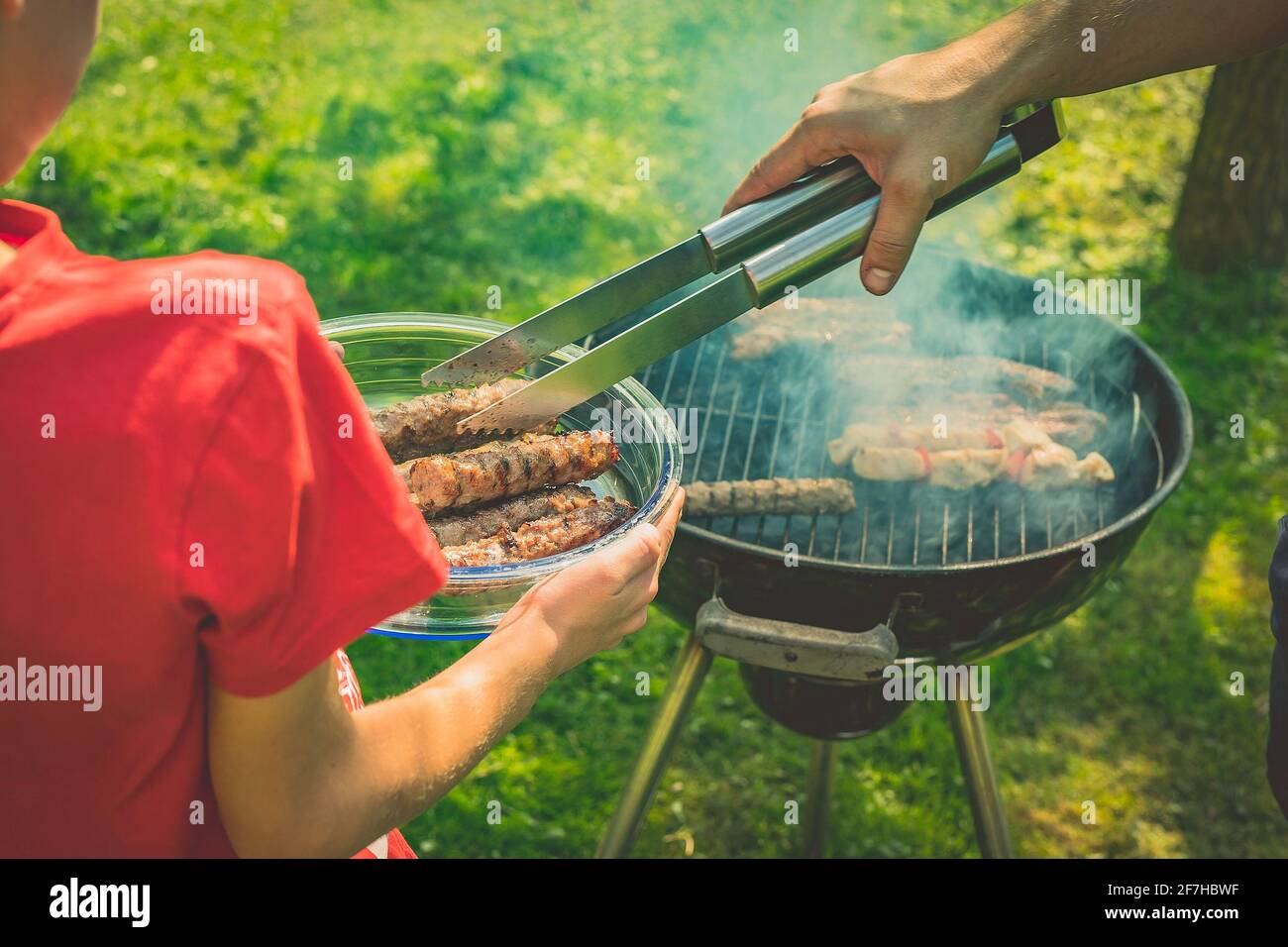 Unknown kid in a red shirt is recieving meat from a grill while holding ...