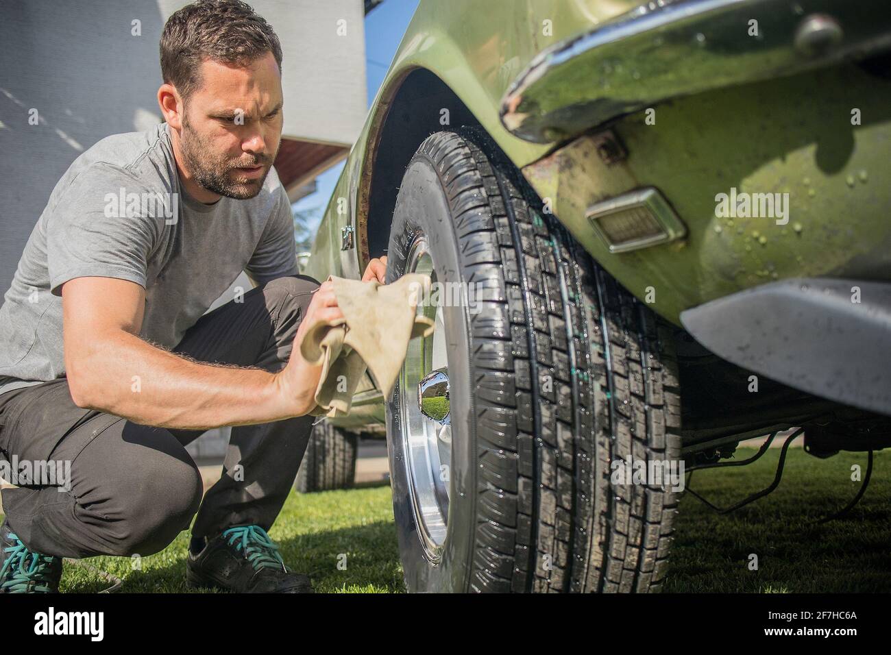 Young man with a hip beard cleaning a wheel of an old green vintage car ...