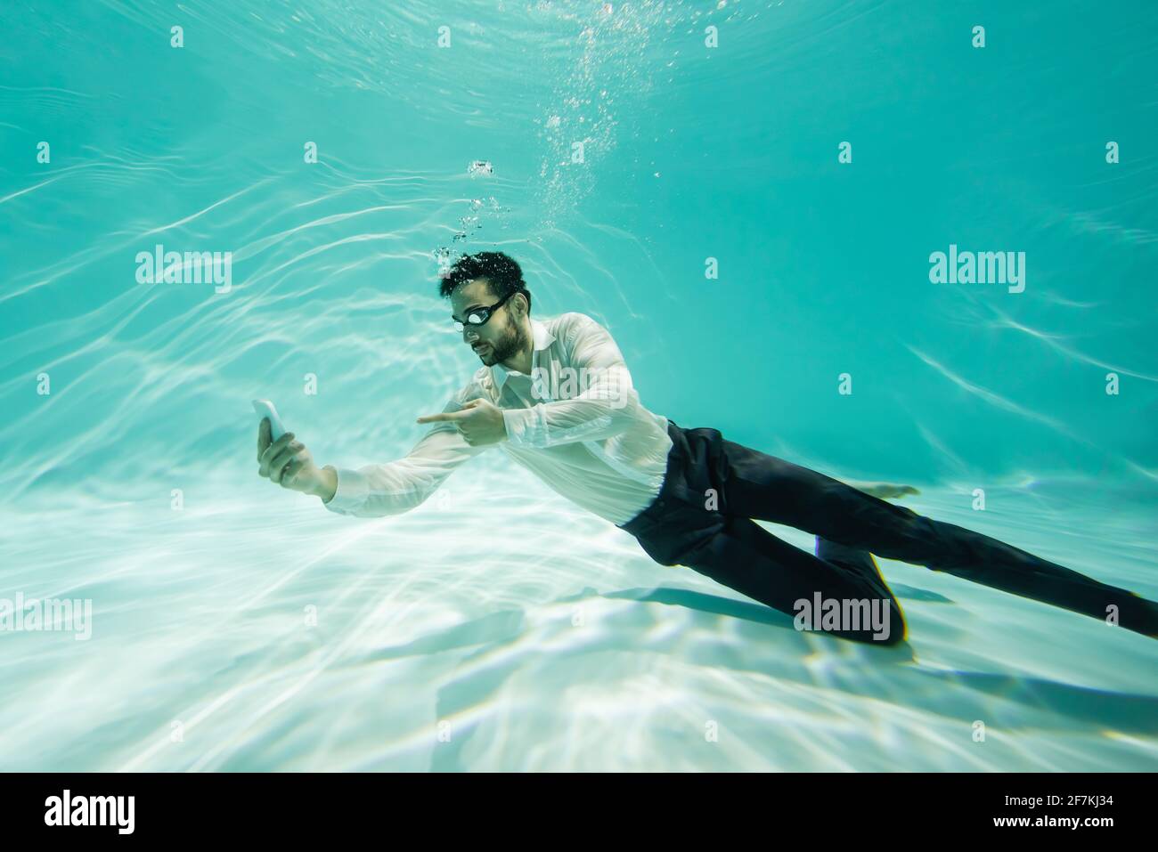Arabian businessman in goggles pointing at smartphone in swimming pool ...