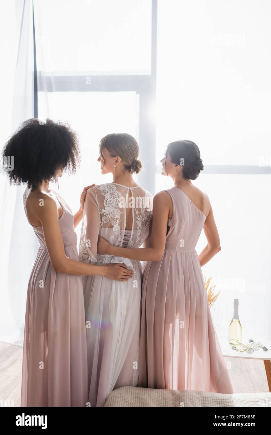 back view of bride near multicultural bridesmaids hugging her in ...