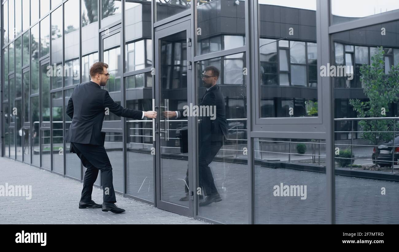full length of businessman in suit opening door in building with glass ...