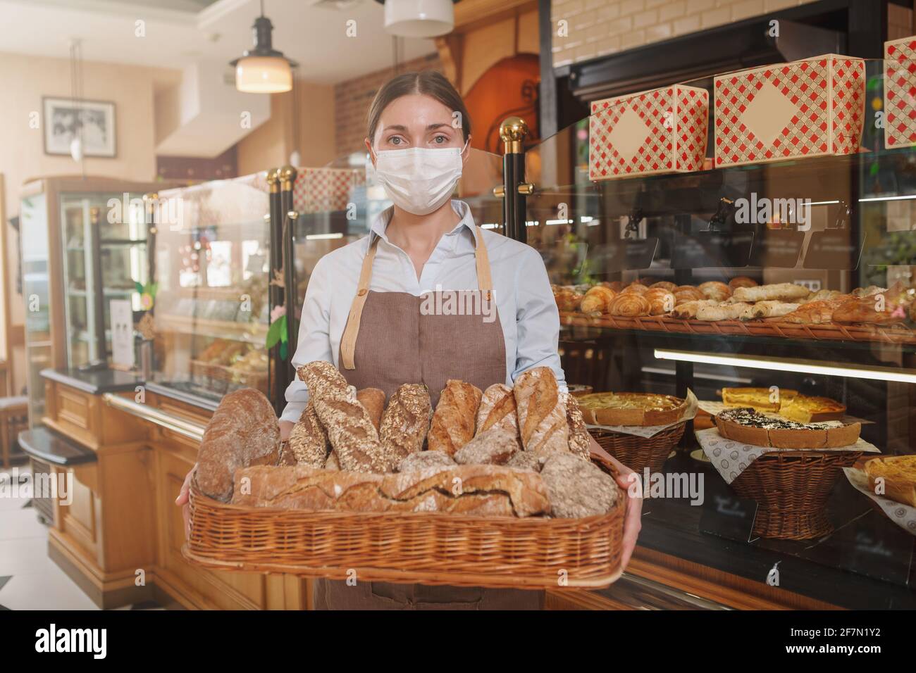Female baker wearing medical face mask, holding basket full of freshly ...