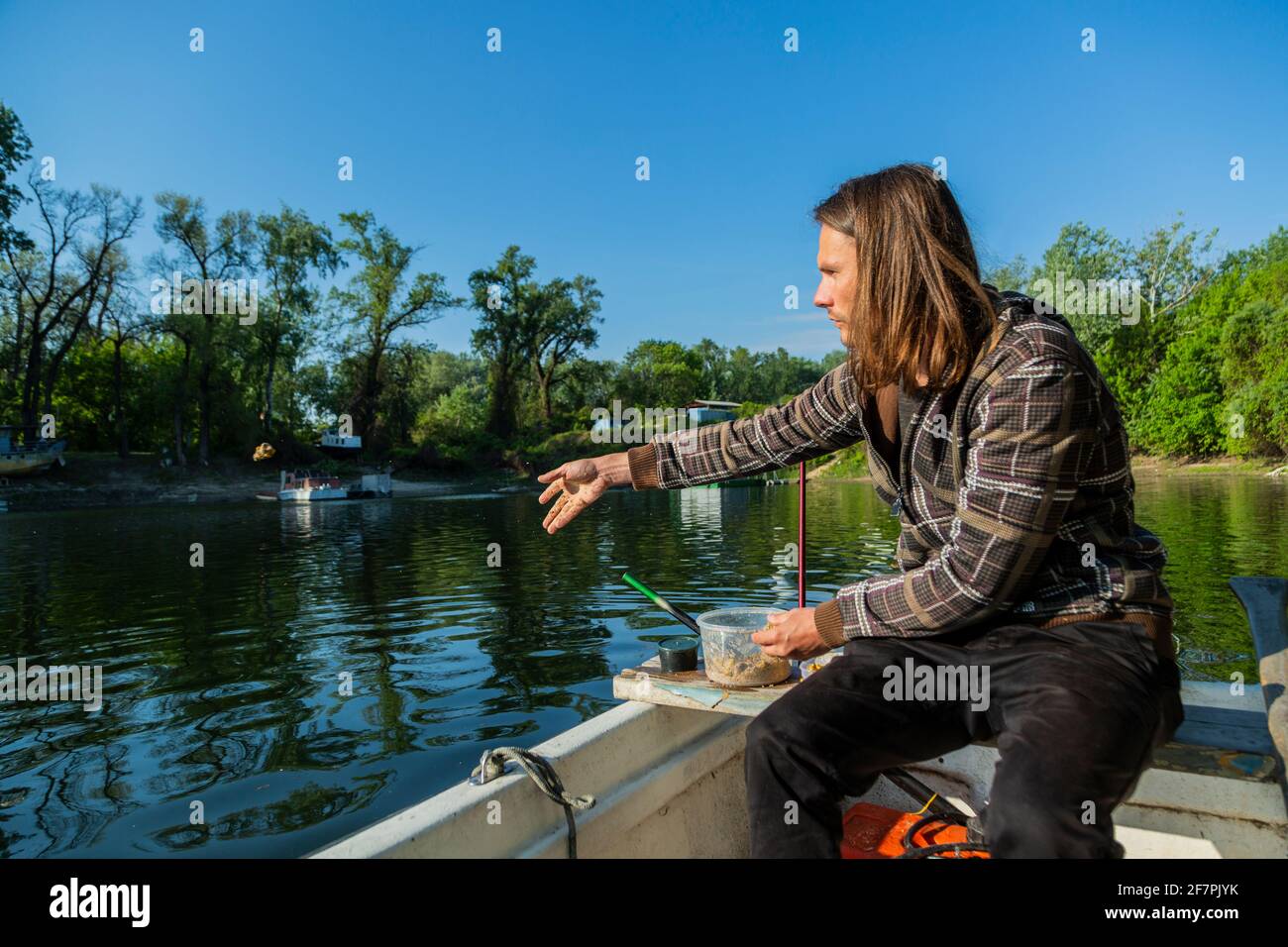 Fisherman throwing chum in lake. He is in white boat with trees around ...