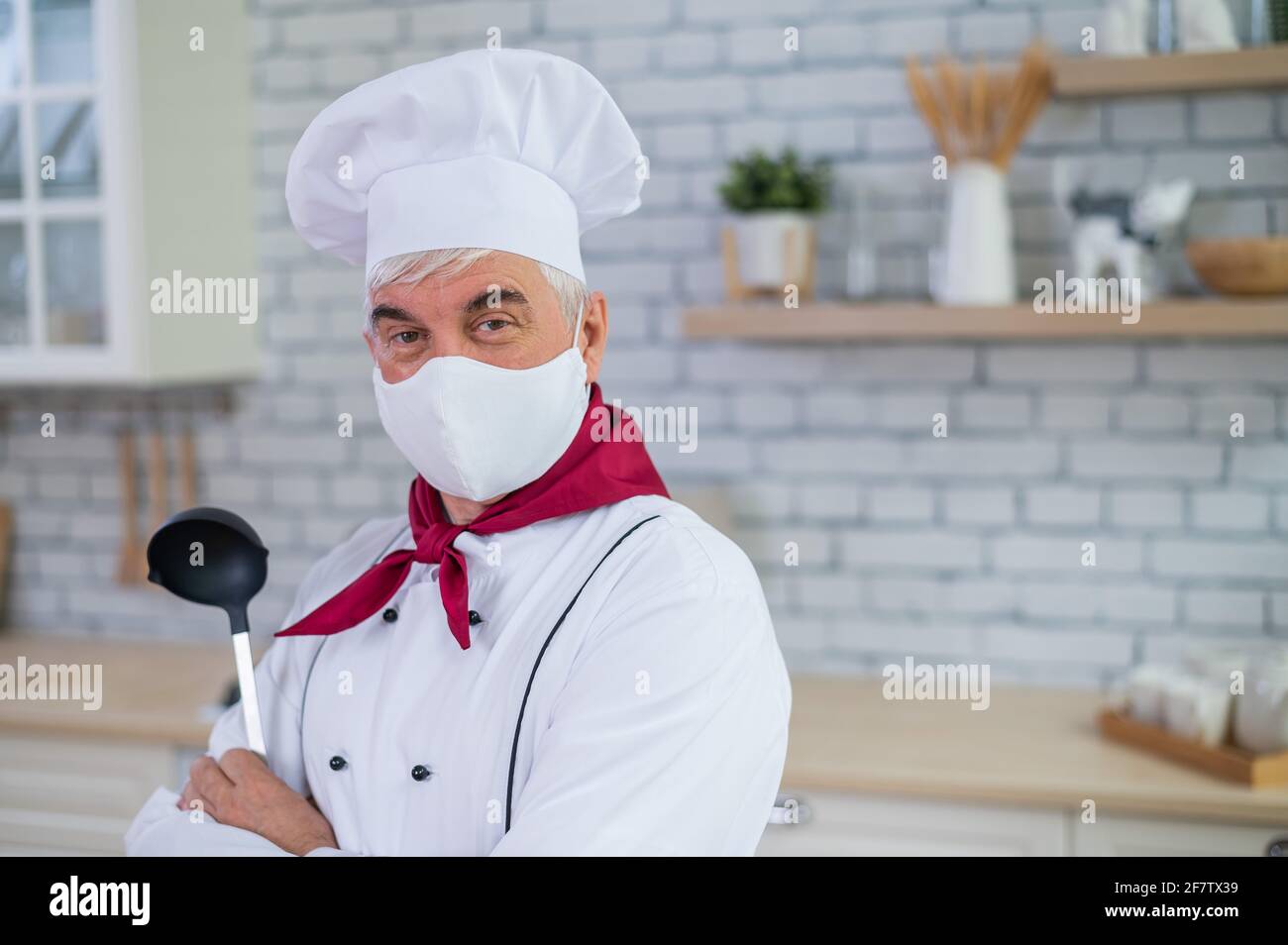 An elderly male chef wearing a mask in the kitchen crossed his arms ...