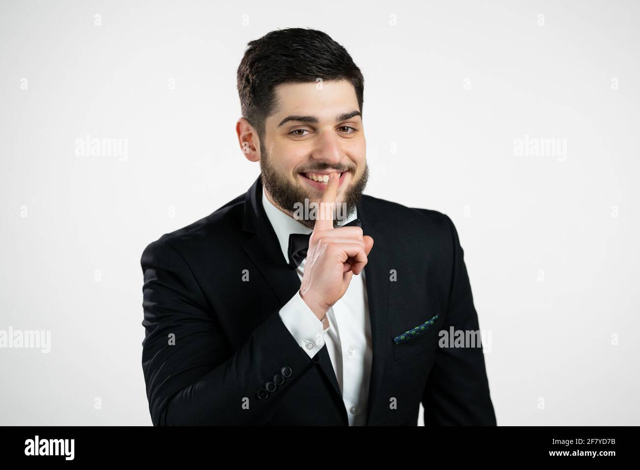Man with beard in tuxedo holding finger on lips over white background ...
