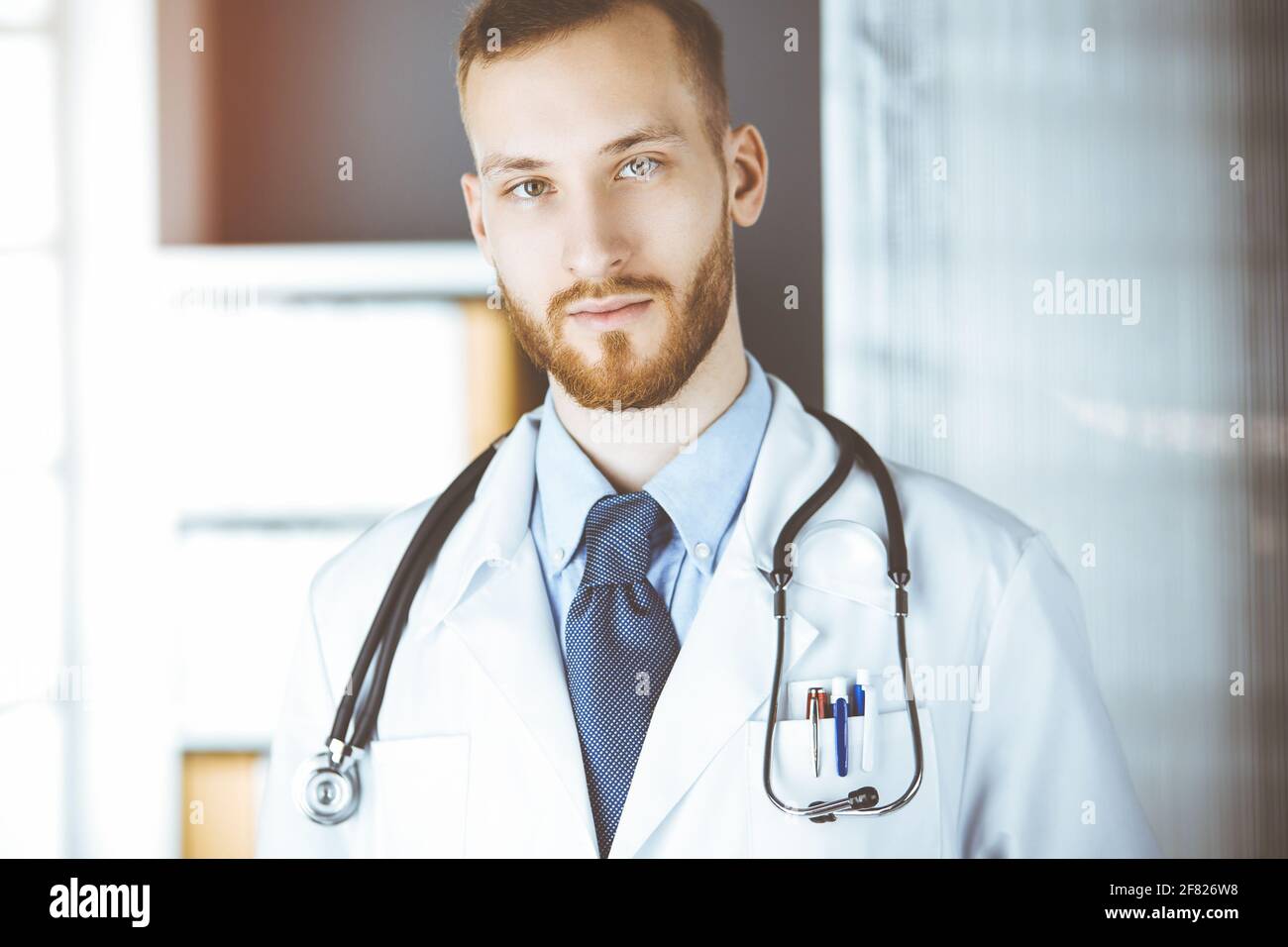 Red-bearded doctor standing straight in sunny clinic near his working ...