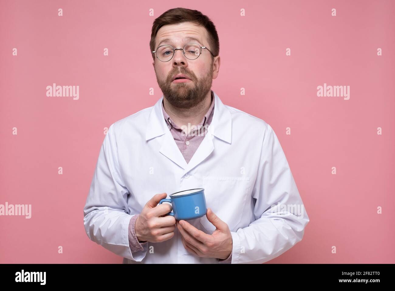 Tired Caucasian doctor holds a mug of coffee in hands, he falls asleep ...