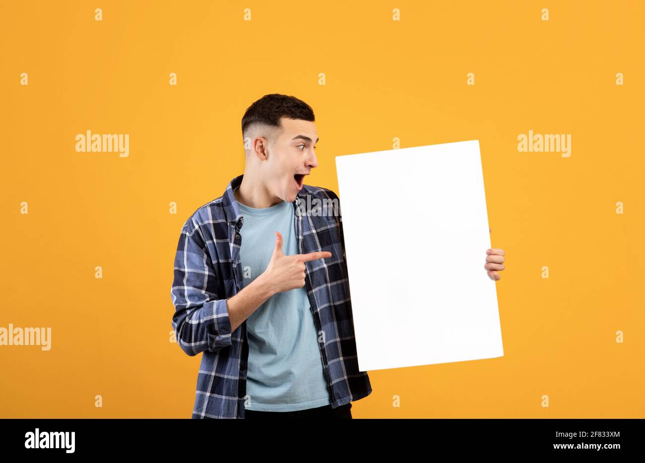 Portrait of shocked young guy pointing at blank paper poster with ...