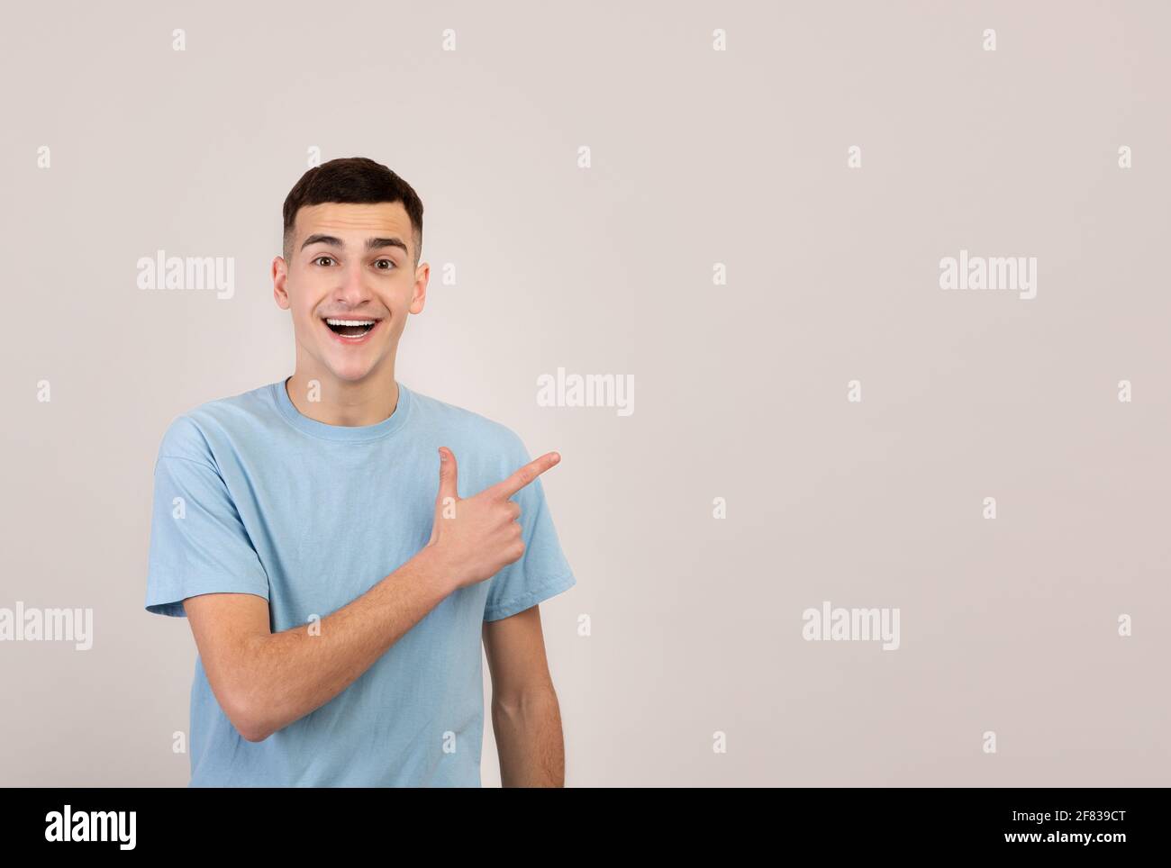 Happy young guy in casual clothes pointing aside at blank space on grey ...