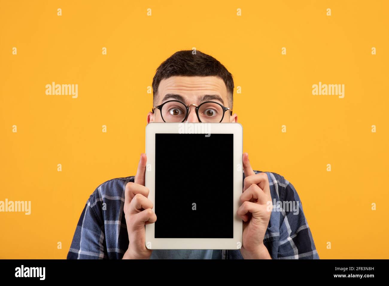Shocked millennial guy hiding behind tablet computer with empty screen ...