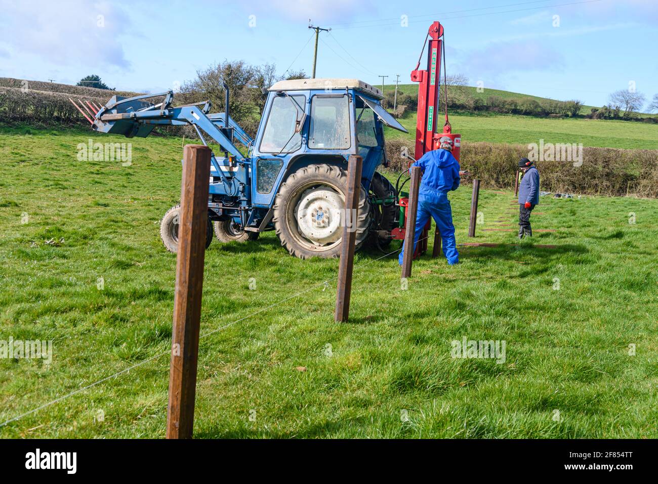 Two farmers men use a hydraulic post driver to install fence posts in a ...