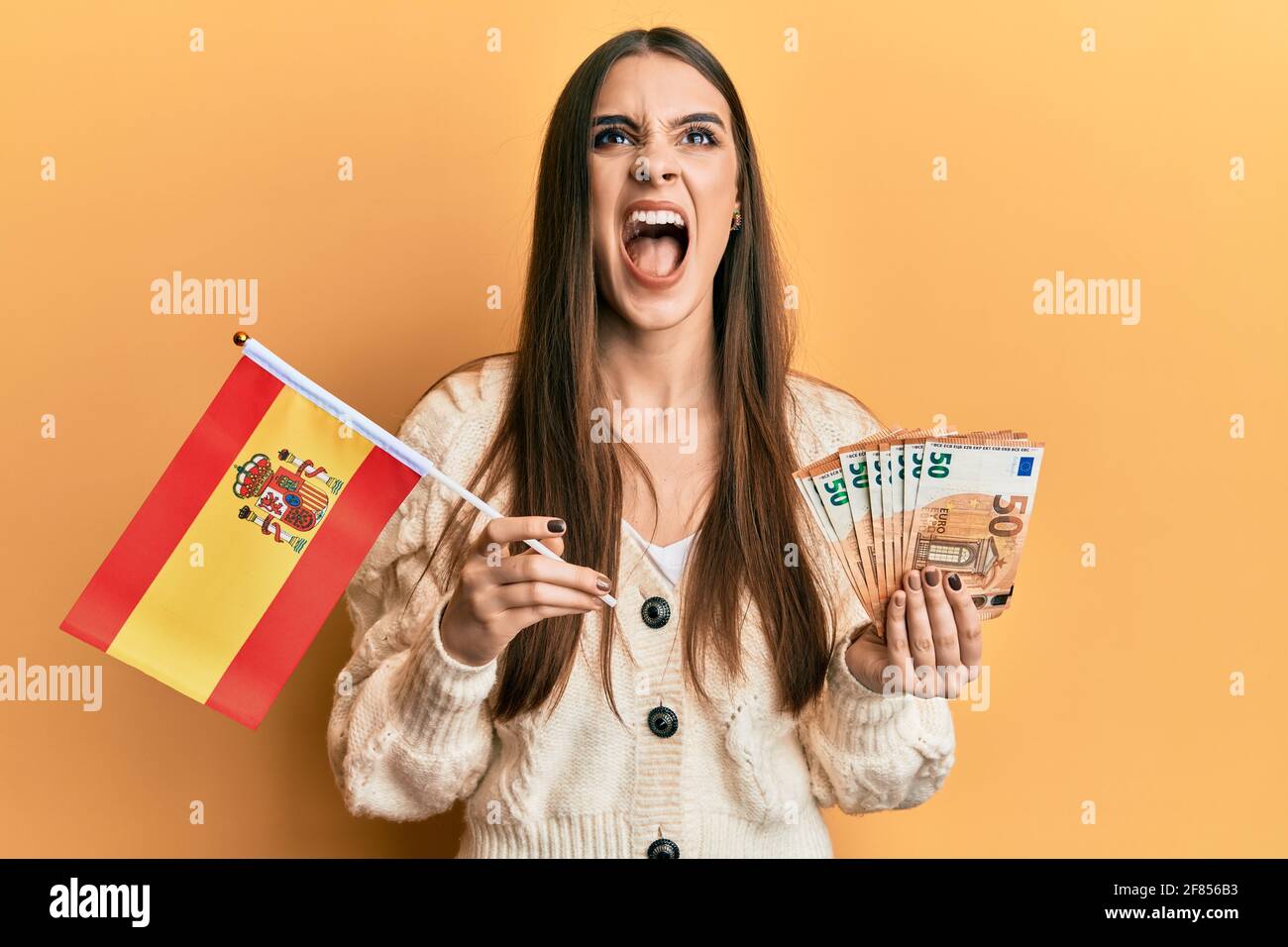 Beautiful brunette young woman holding spain flag and euros banknotes ...