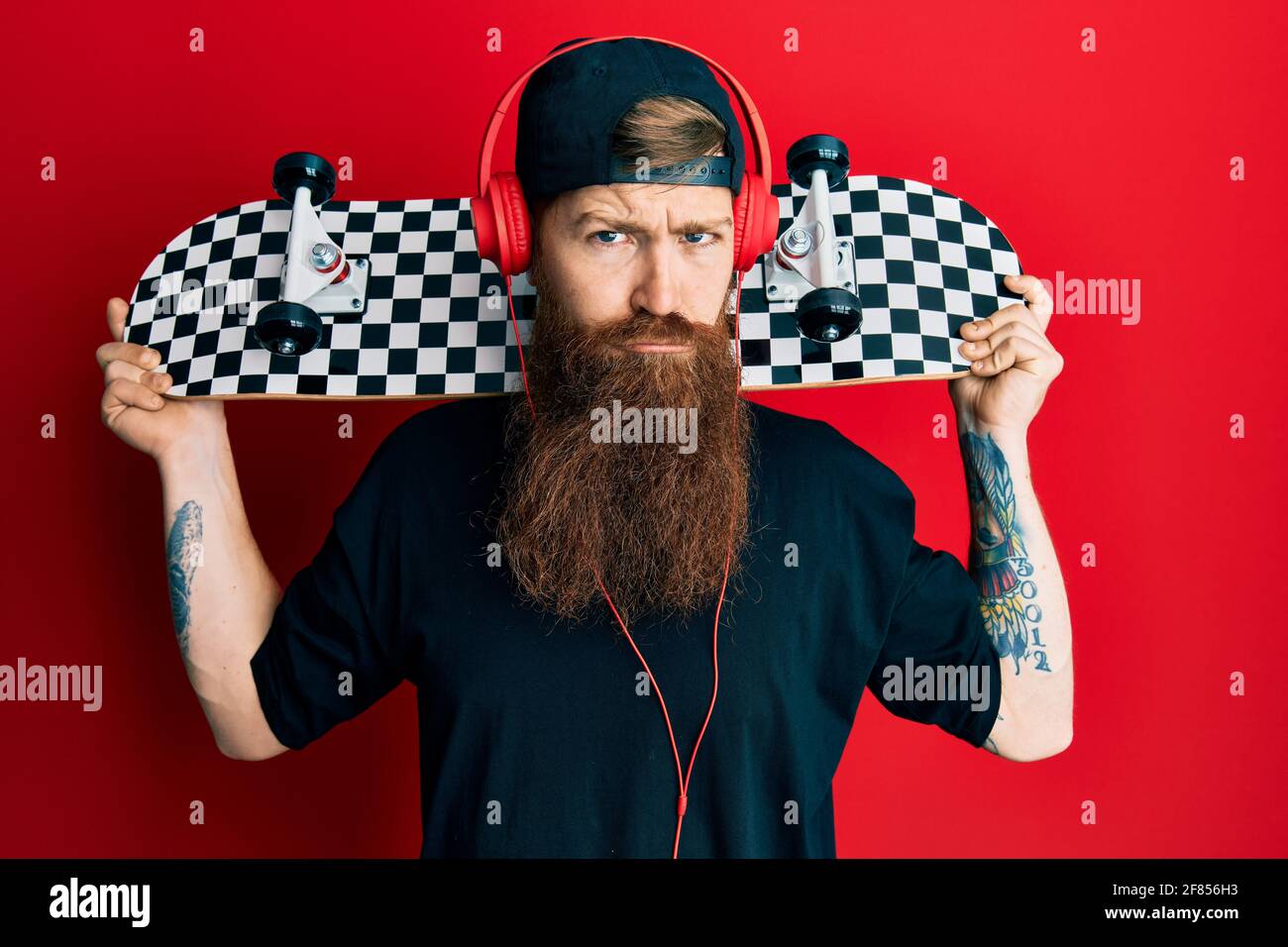 Redhead man with long beard holding skate wearing headphones depressed ...