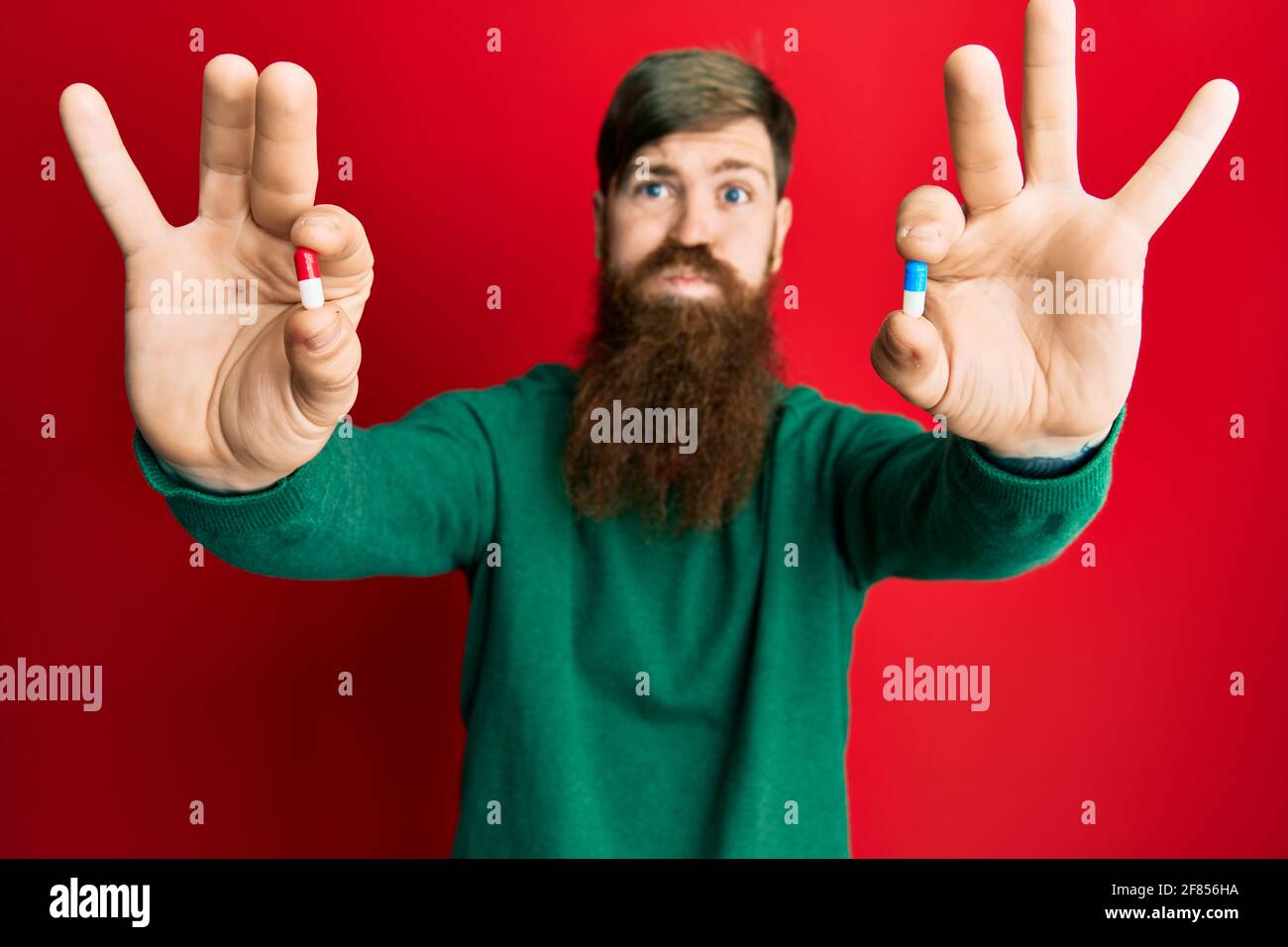 Redhead man with long beard holding two different pills puffing cheeks ...