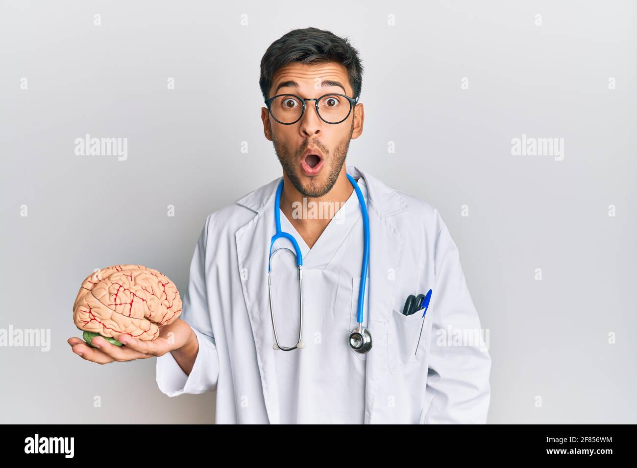 Young handsome man wearing doctor coat holding brain scared and amazed ...