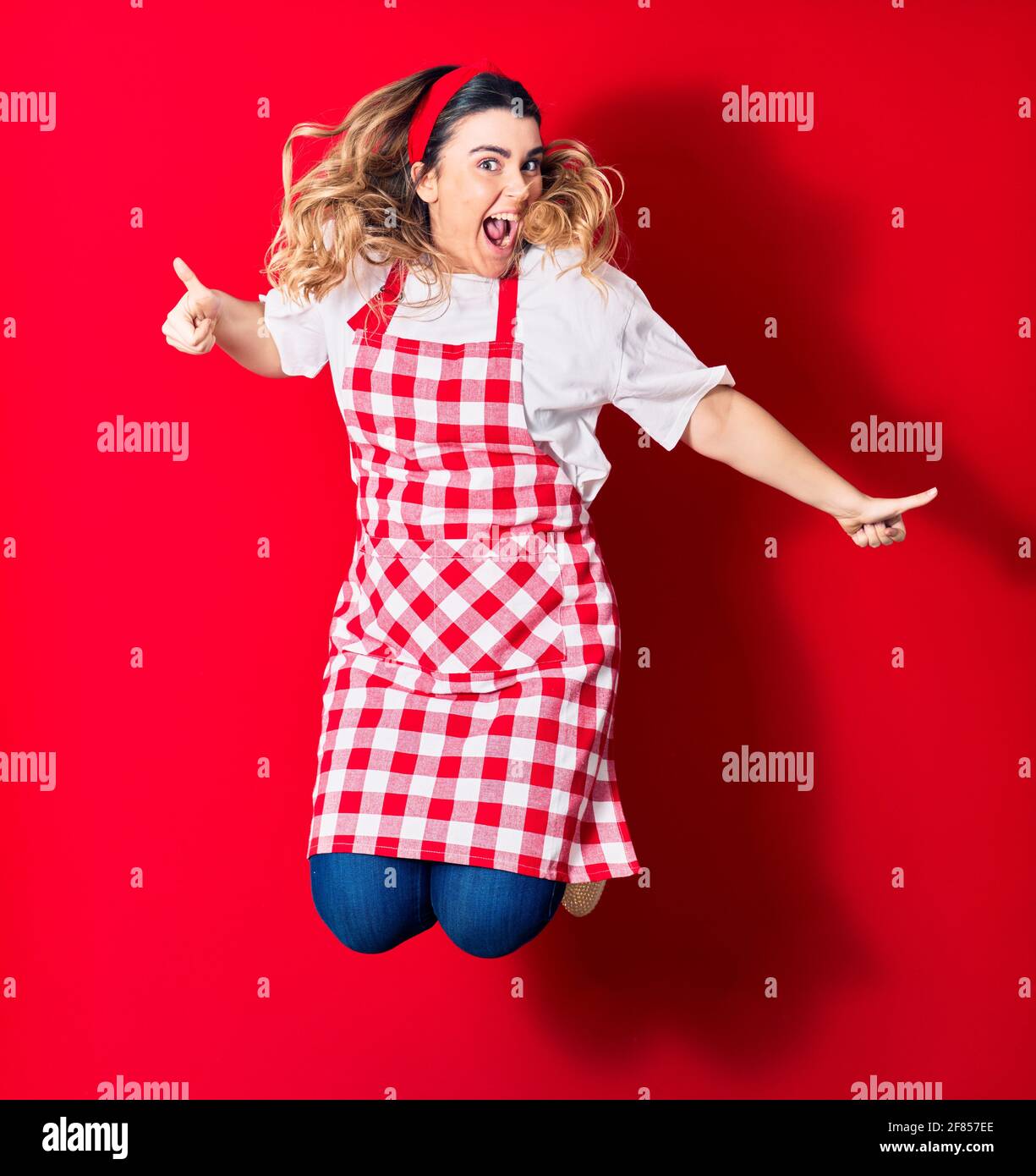 Young beautiful blonde cook woman wearing apron smiling happy. Jumping ...