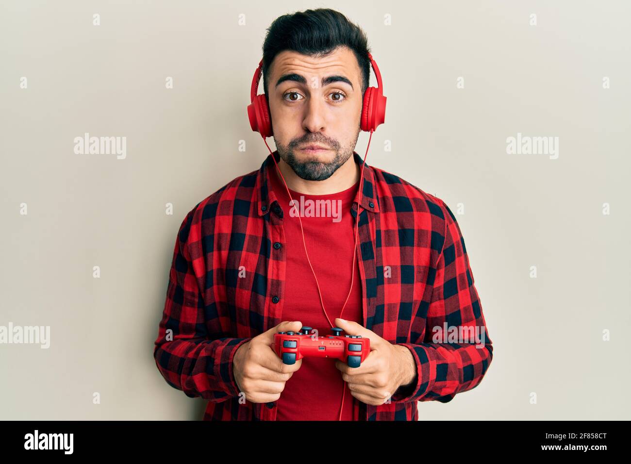 Young hispanic man playing video game holding controller puffing cheeks ...