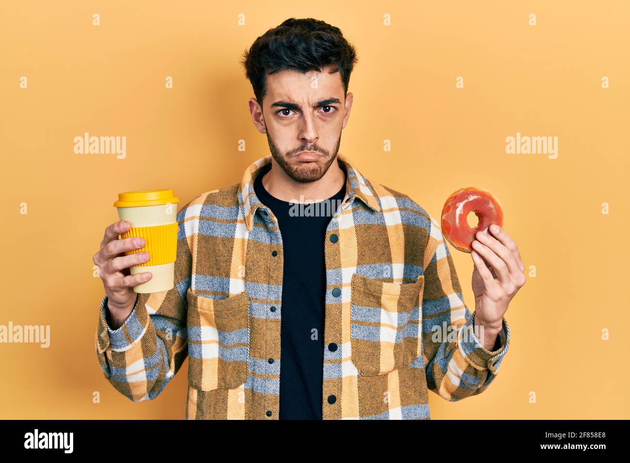 Young hispanic man eating doughnut and drinking coffee depressed and ...