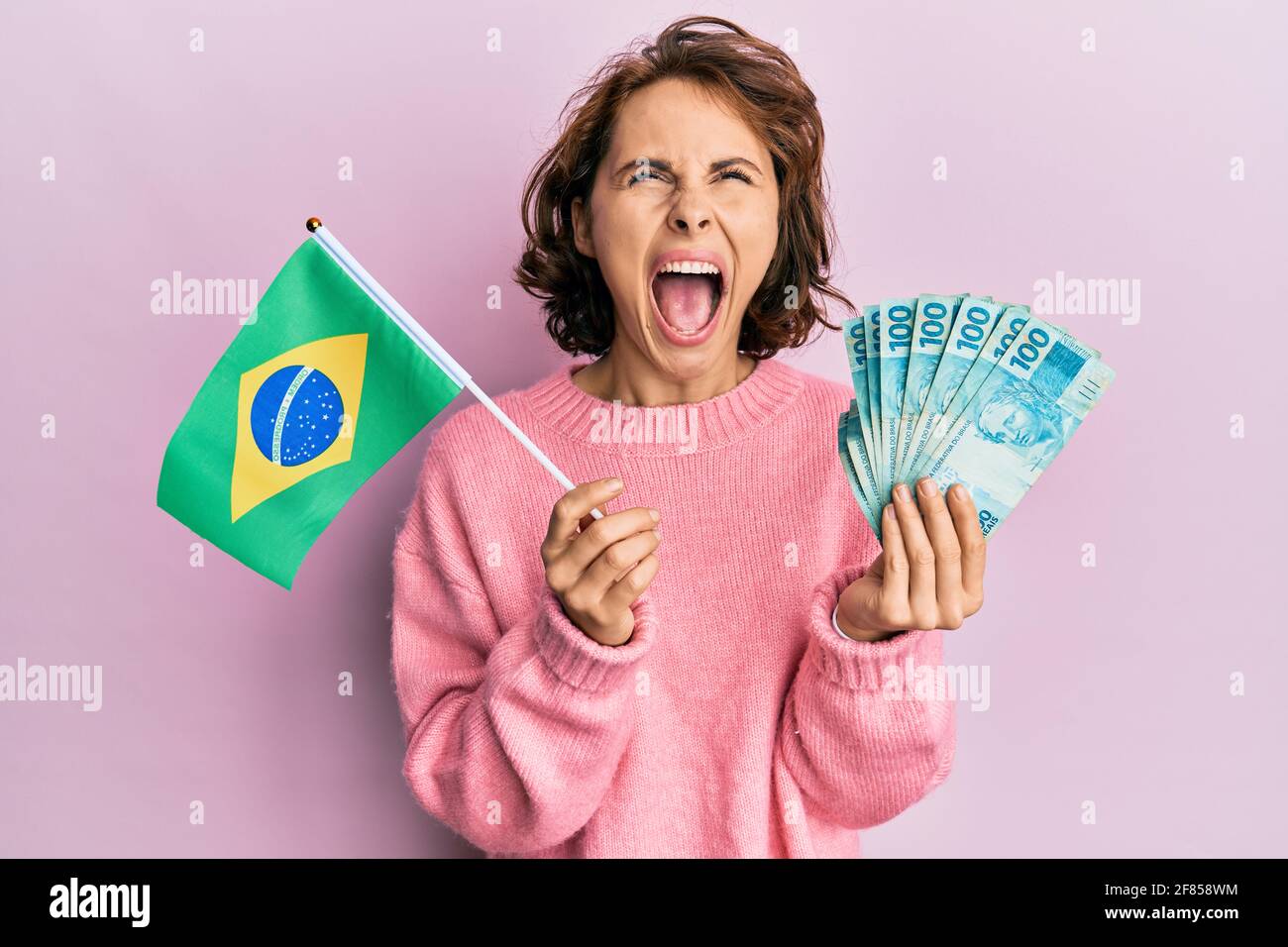 Young brunette woman holding brazil flag and real banknotes angry and ...