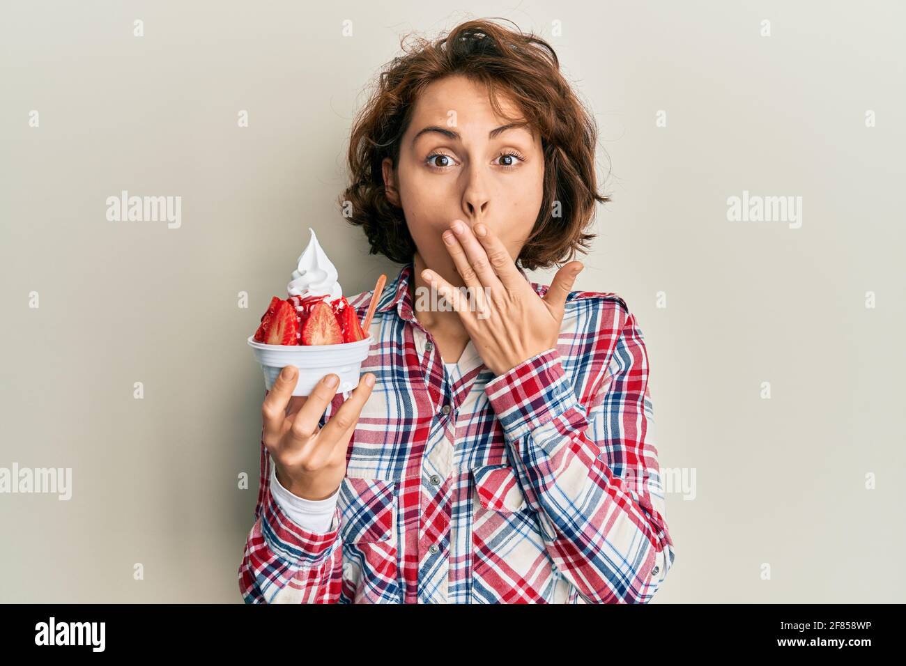 Young brunette woman eating strawberry ice cream covering mouth with ...