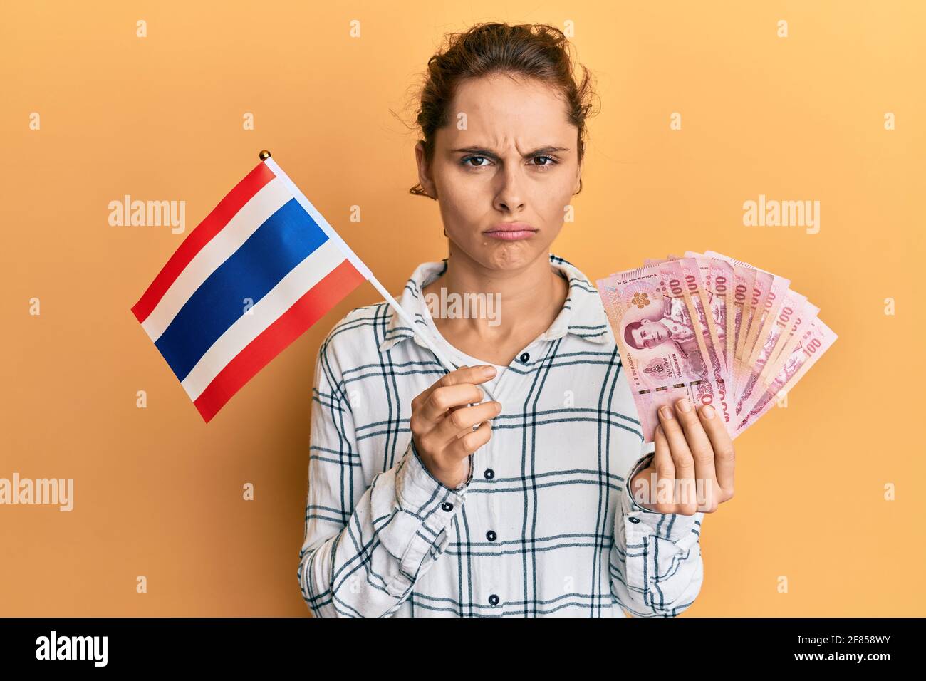 Young brunette woman holding thailand flag and baht banknotes depressed ...