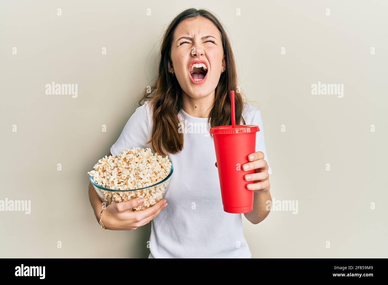 Young brunette woman eating popcorn and drinking soda angry and mad ...