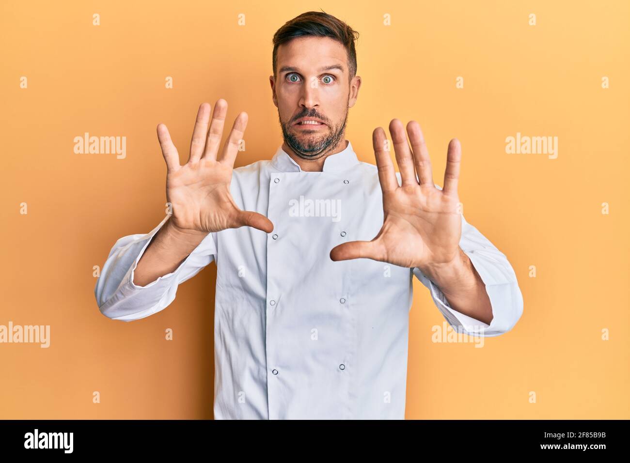 Handsome man with beard wearing professional cook uniform afraid and ...