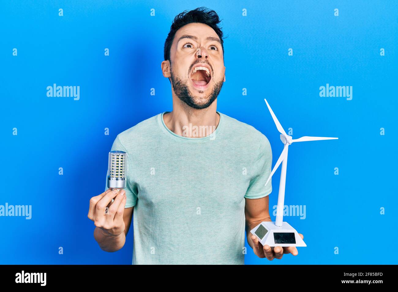 Young hispanic man holding windmill and led lightbulb angry and mad ...