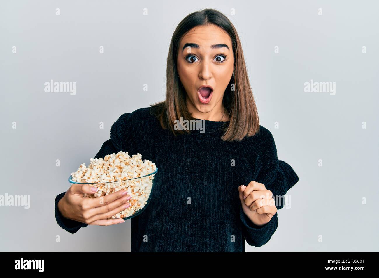Young brunette girl eating popcorn scared and amazed with open mouth for surprise, disbelief ...
