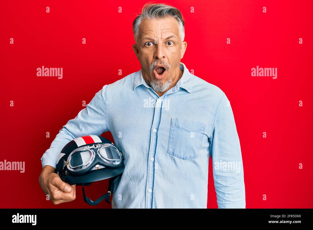 Middle age grey-haired man holding motorcycle helmet scared and amazed ...