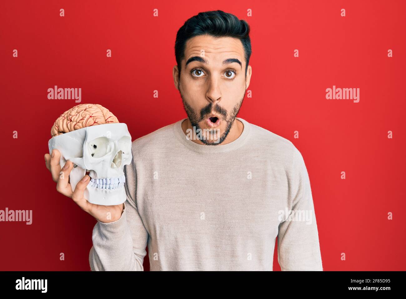 Young hispanic man holding skull with brain scared and amazed with open ...