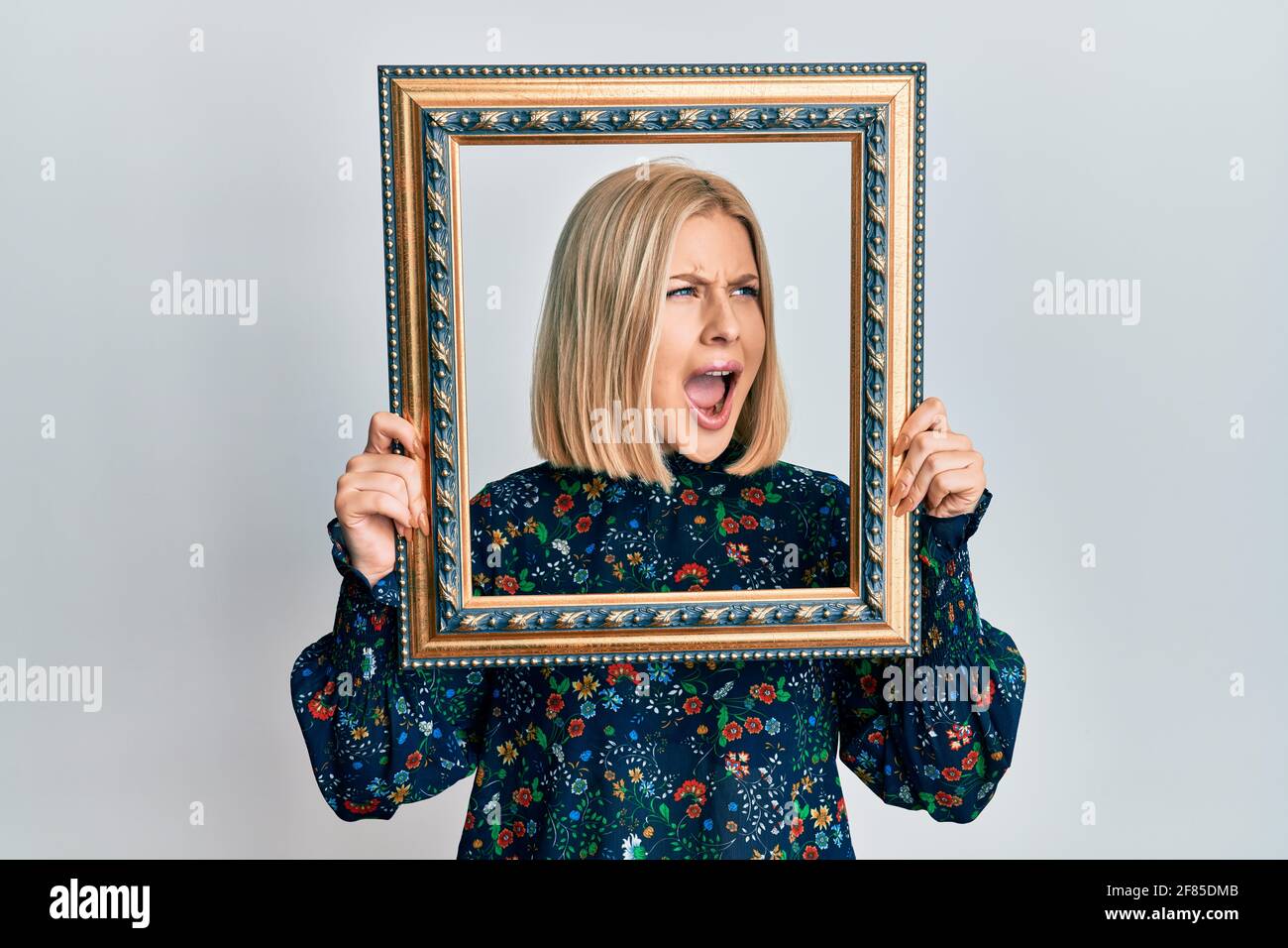 Young blonde woman holding empty frame angry and mad screaming ...