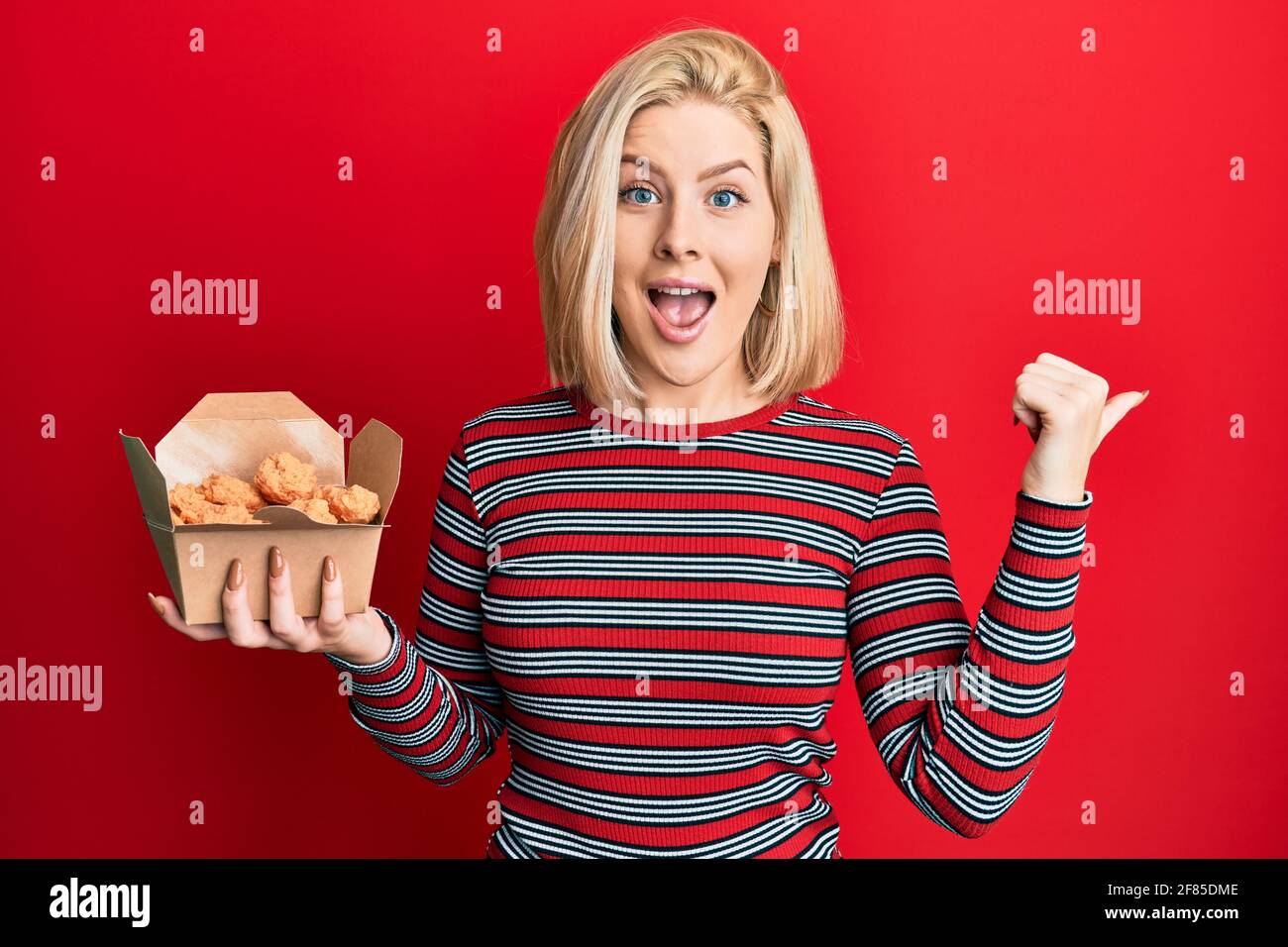 Young blonde woman eating fried chicken pointing thumb up to the side ...