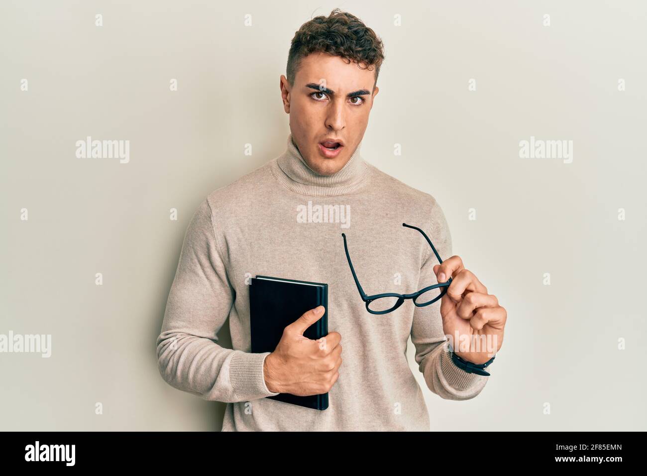 Hispanic young man reading a book and holding glasses in shock face ...
