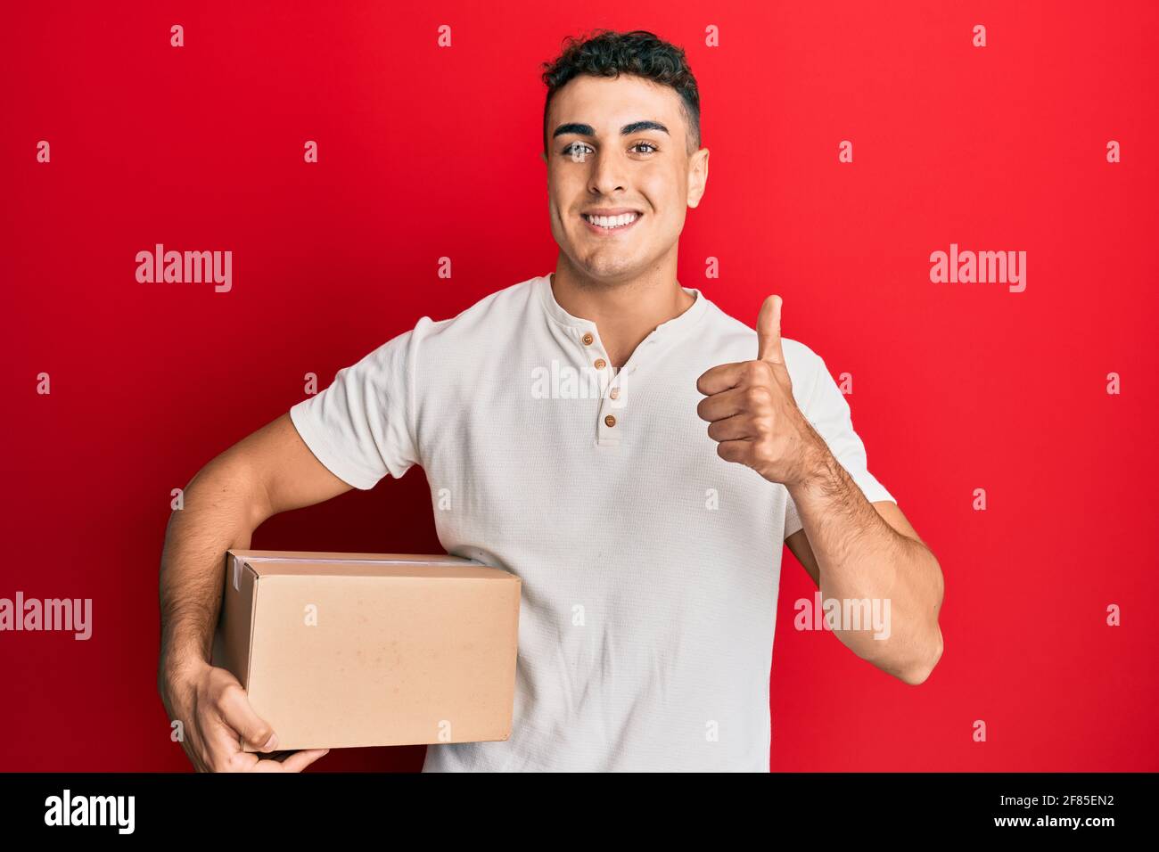 Hispanic young man holding delivery package smiling happy and positive ...
