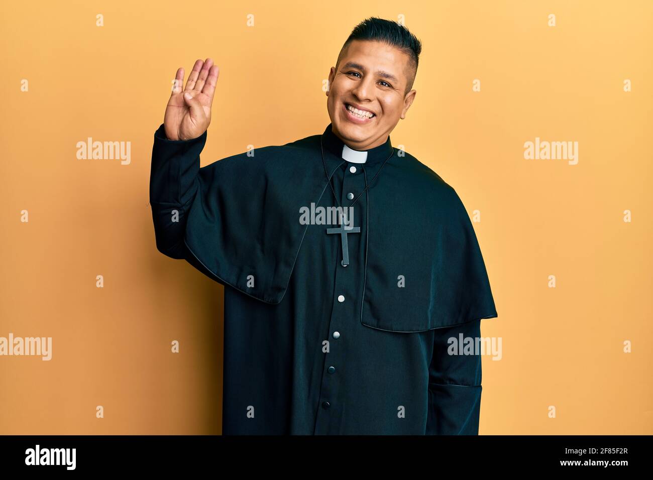 Young latin priest man standing over yellow background waiving saying ...